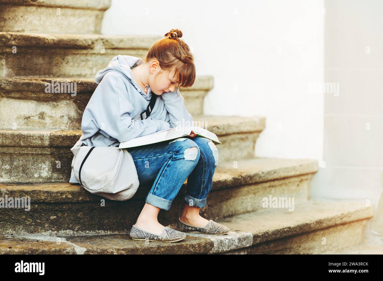 Kid girl reading book outside, sitting on stairs, wearing grey ...
