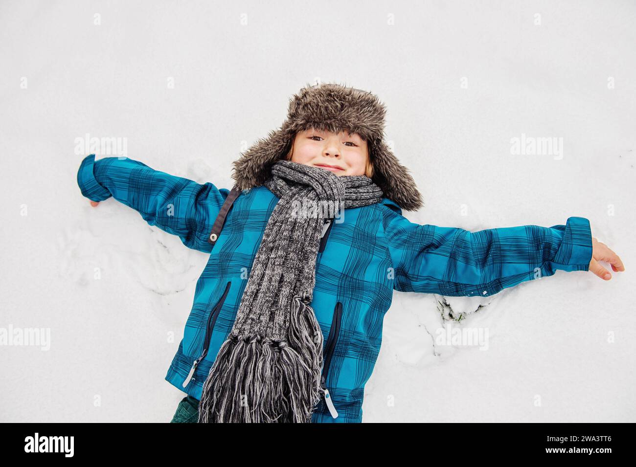 Little boy lying on white fresh snow, wearing warm hat, scarf and blue ...