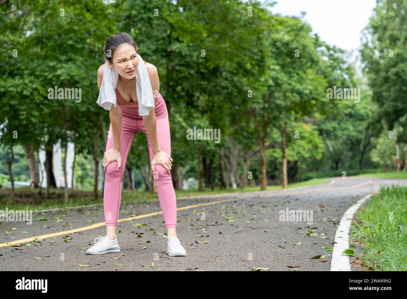 Tired young Asian woman in fitness clothes stops and leans down to take ...