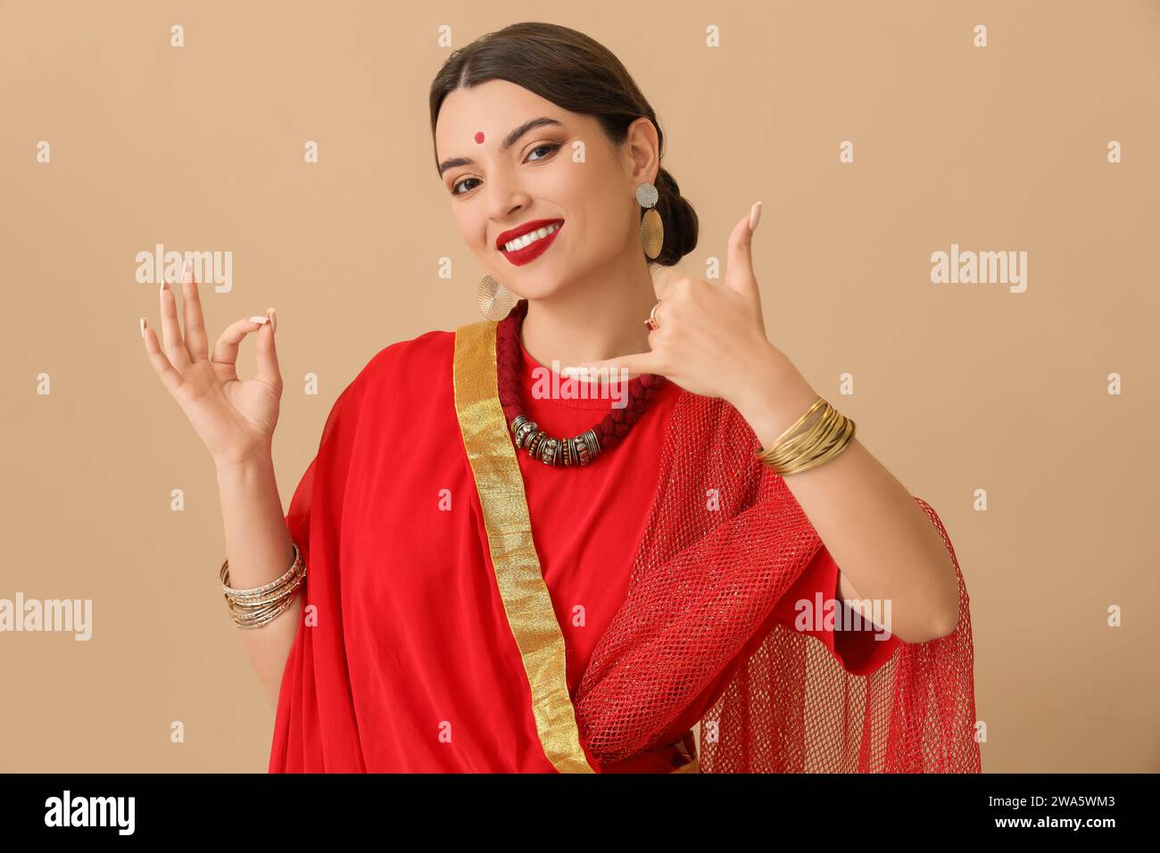 Beautiful young Indian woman showing "call me" and OK gesture on beige ...