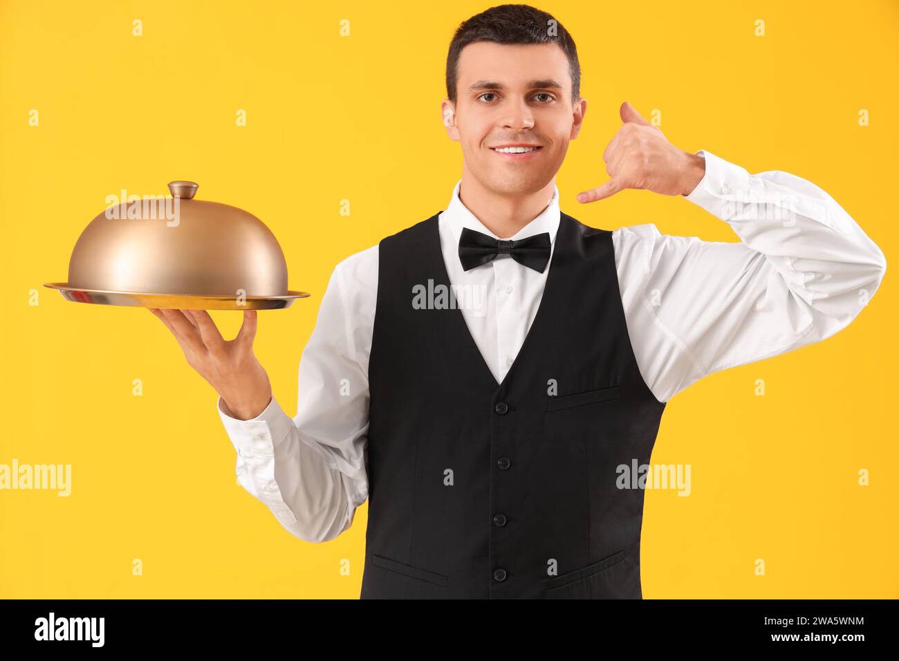 Male waiter with cloche showing "call me" gesture on yellow background ...