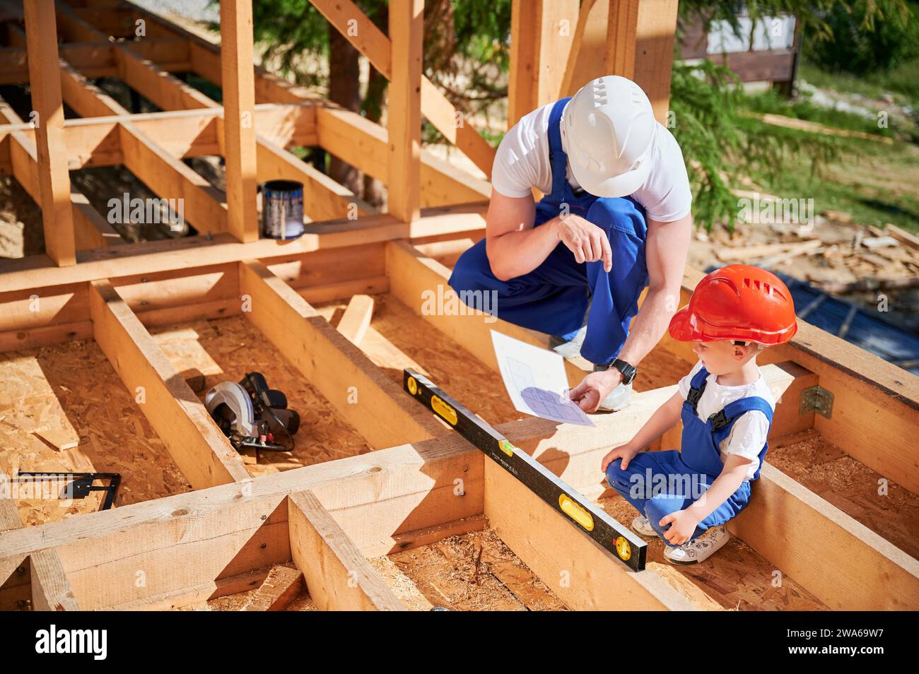 Father with toddler son constructing wooden frame house. Male builder showing his son the ...