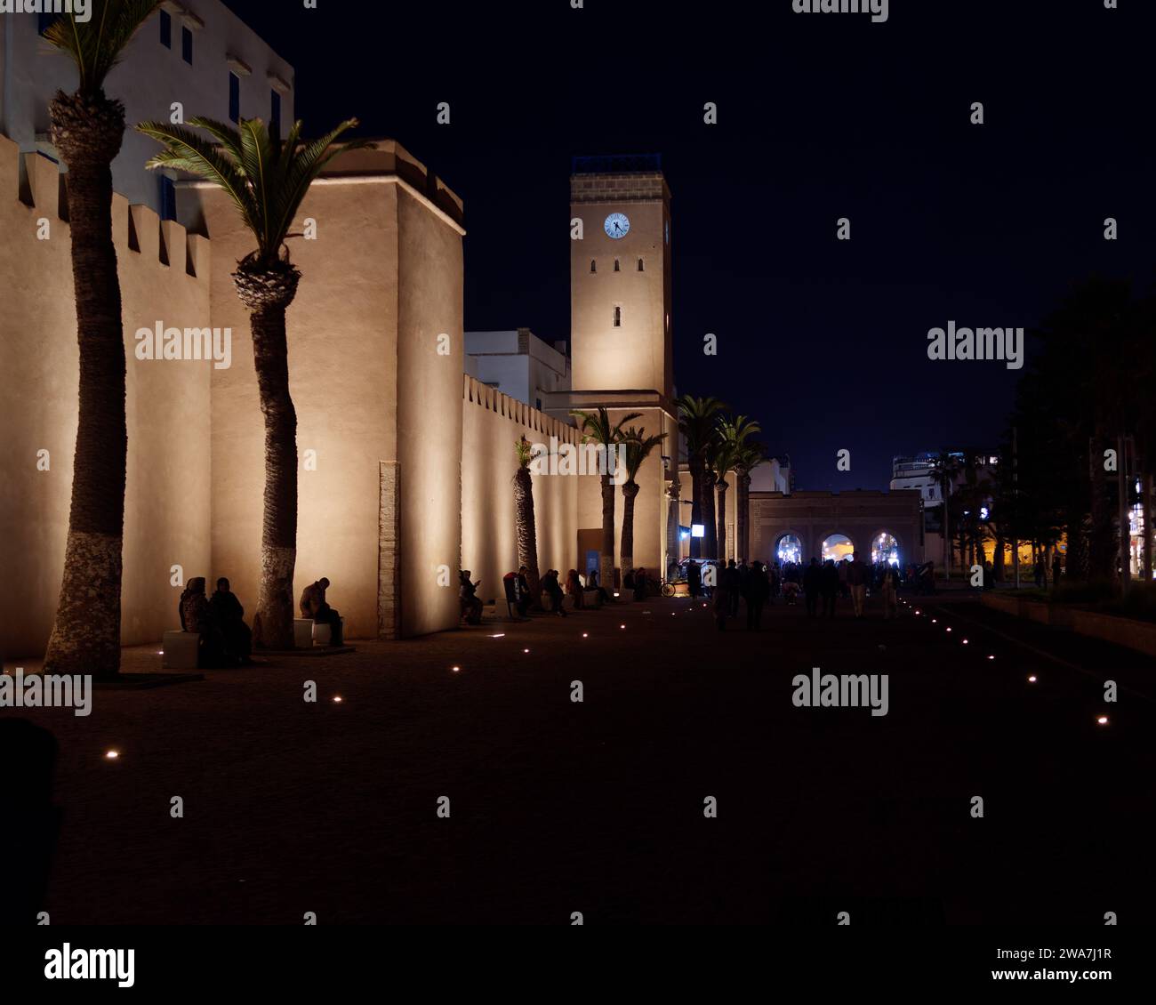 People sit on benches at night beside floodlit city walls with clock ...