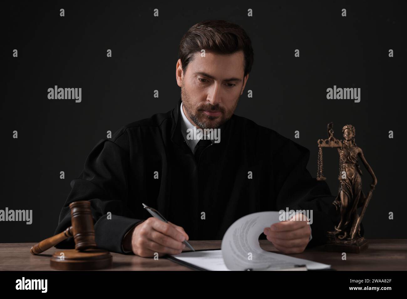 Judge with gavel writing in papers at wooden table against black ...