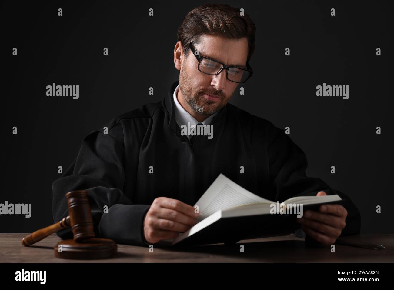 Judge with gavel reading book at wooden table against black background ...