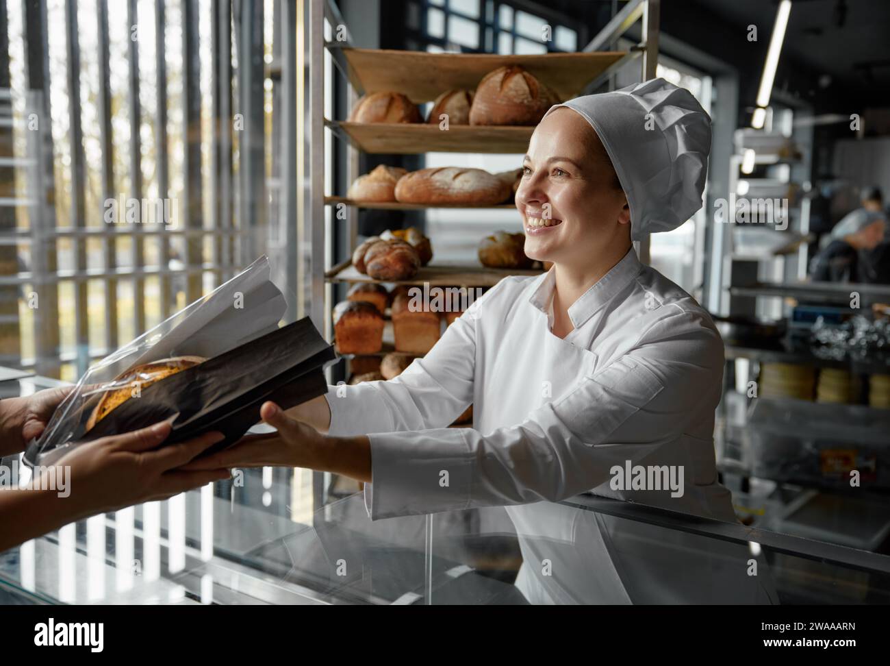 Friendly smiling saleswoman in bakery passing bread over counter desk ...