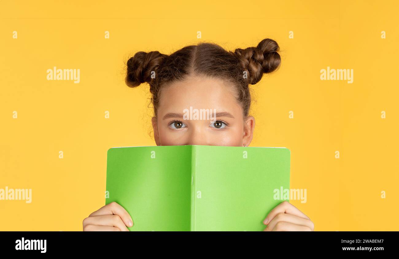 Curious young girl with buns hairstyle peeks over a green book, her ...