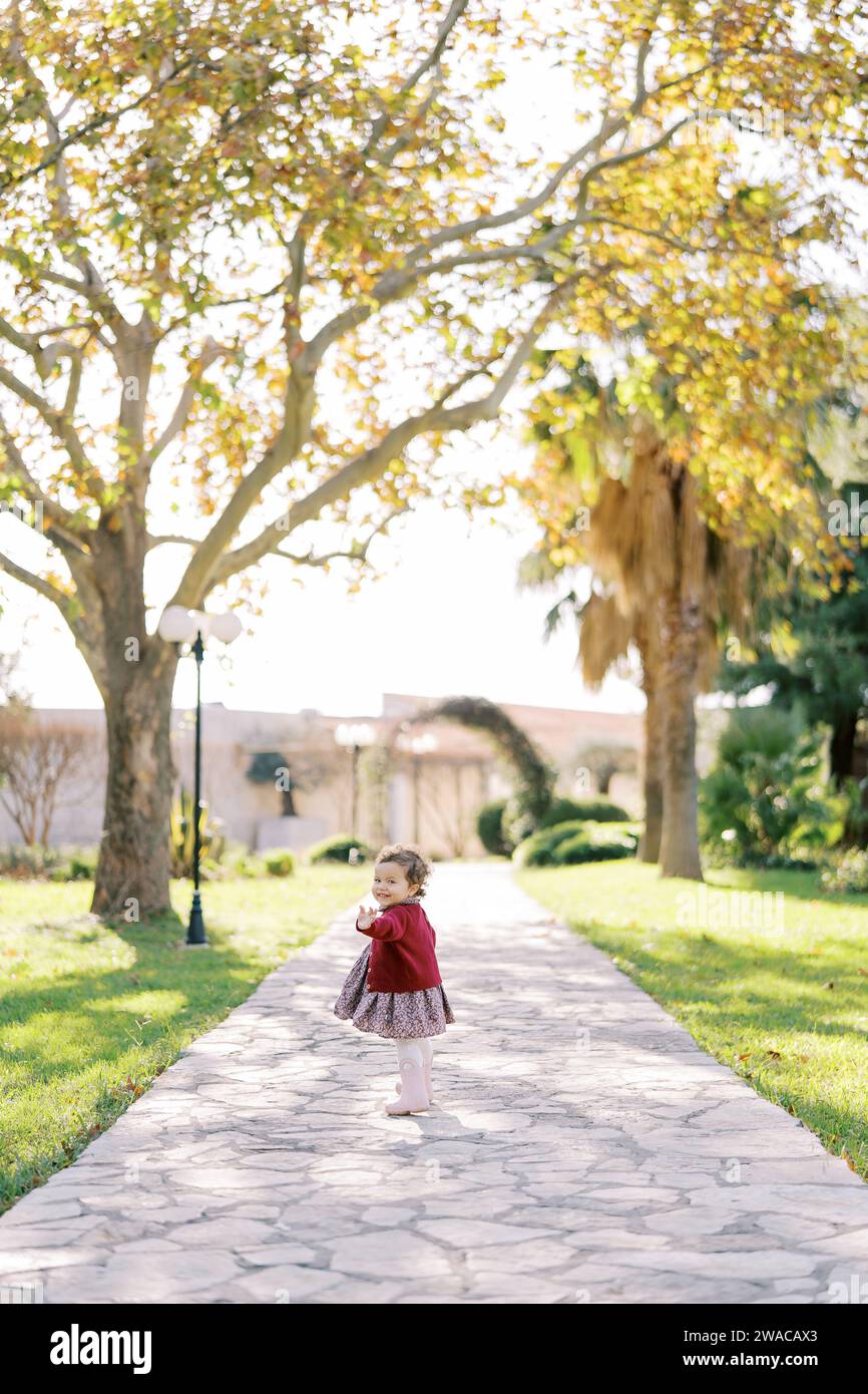 Little girl stands half-turned on a paved path in the park and waves ...