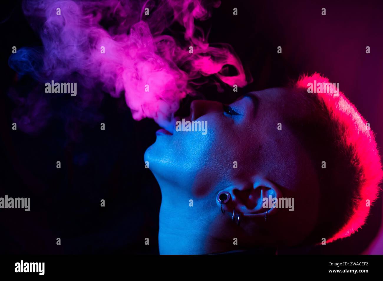 Portrait asian woman with shaved temples smokes vape in neon light ...