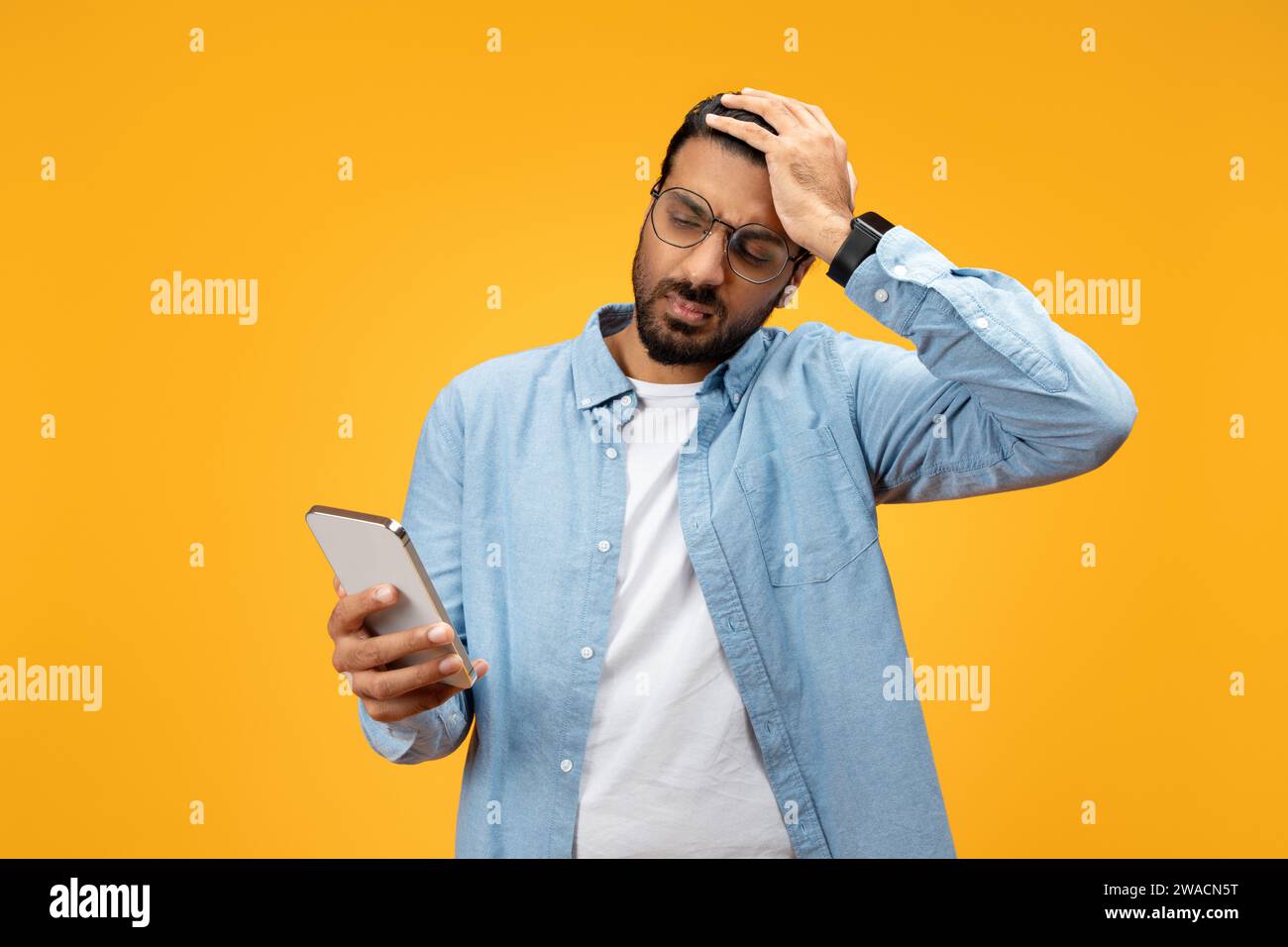 A perplexed man in a blue shirt holds his smartphone and looks at it ...
