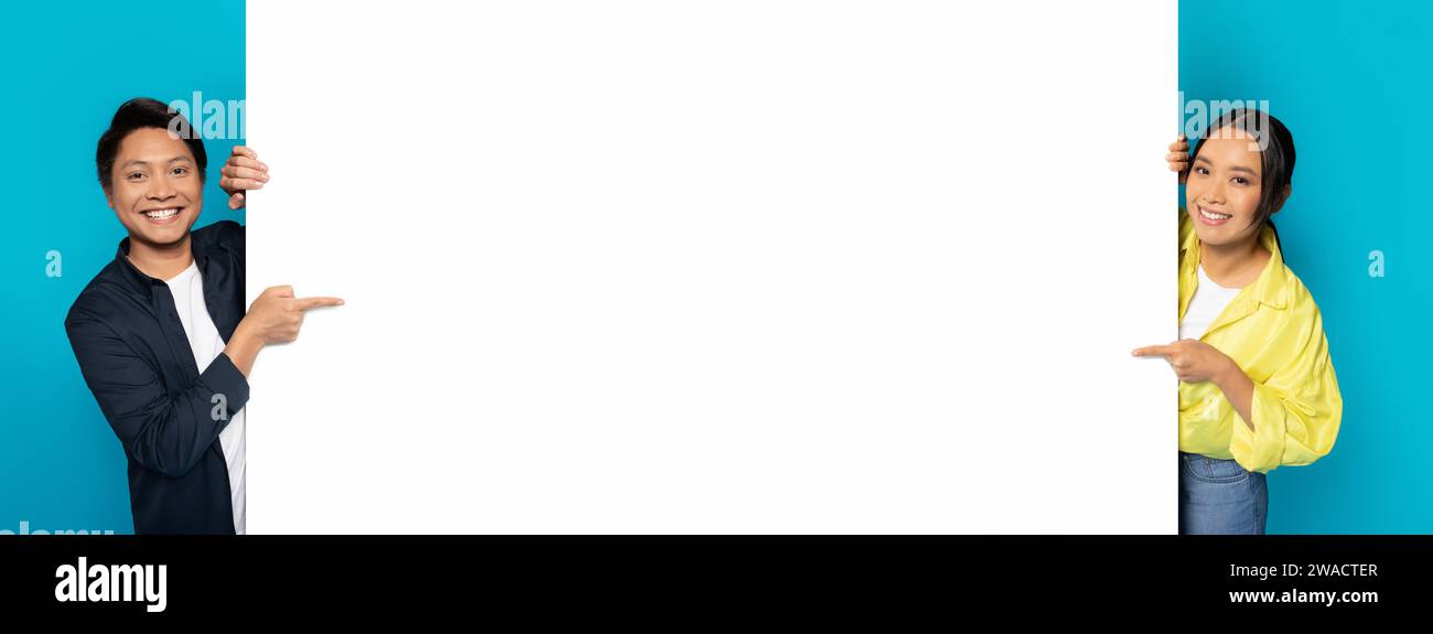 Asian man and woman joyfully pointing at a blank wide white banner ...
