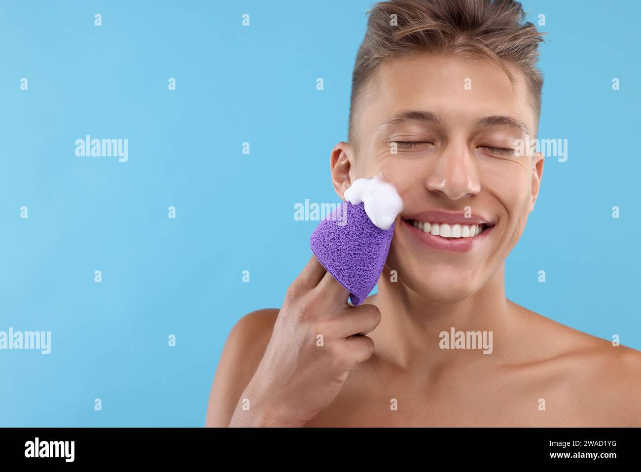 Happy young man washing his face with sponge on light blue background. Space for text Stock ...