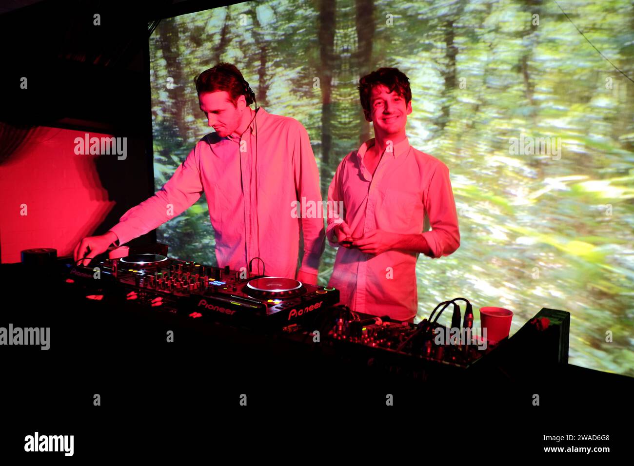 Two DJs behind a mixing desk in pink light, a green forest projected ...