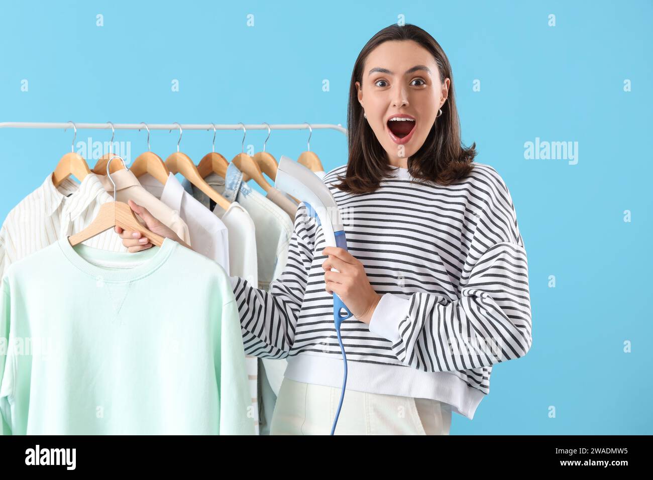 Surprised young woman with modern steamer and clothes on blue ...
