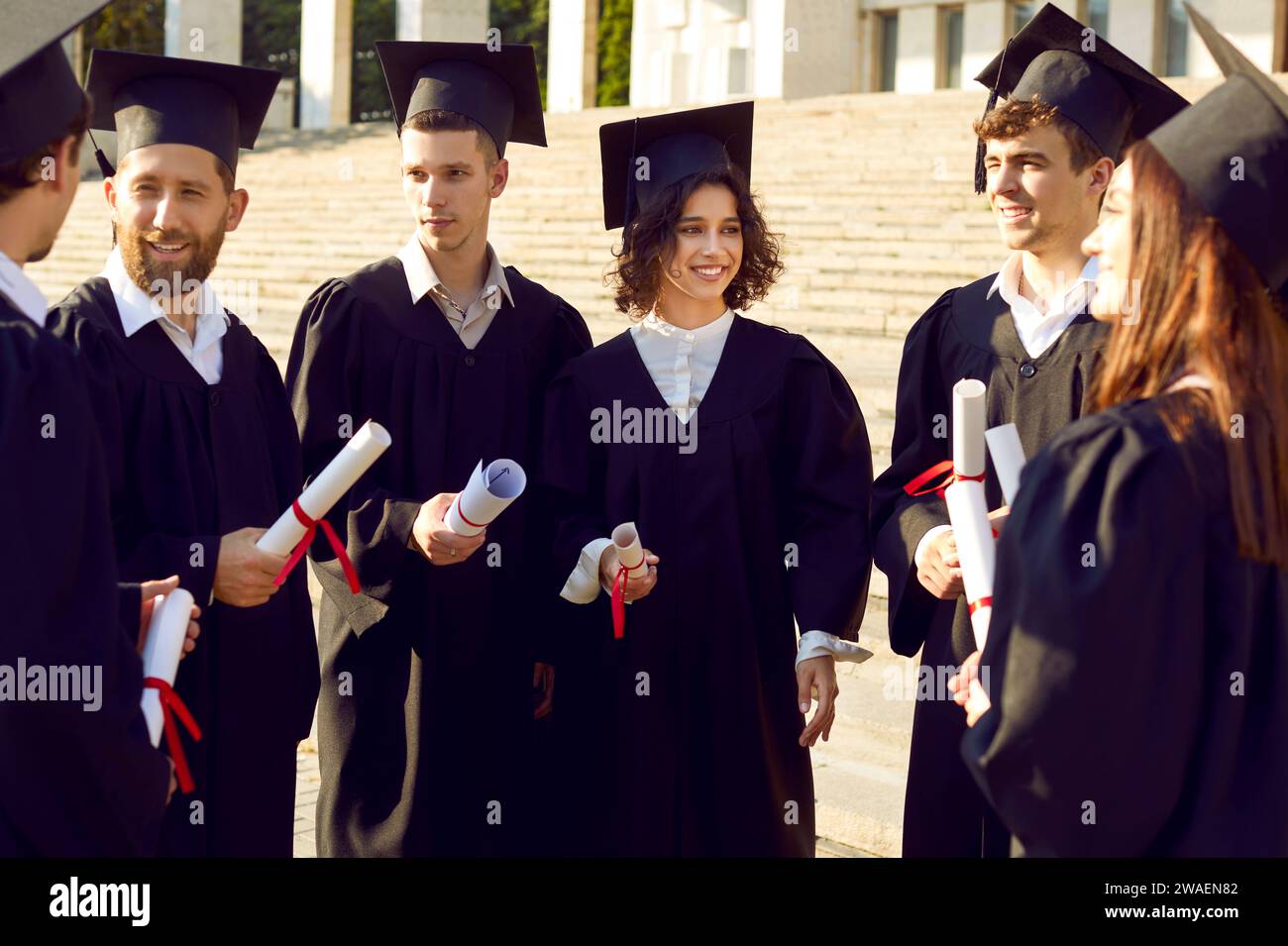 Happy graduate students in black graduation robes talking near ...