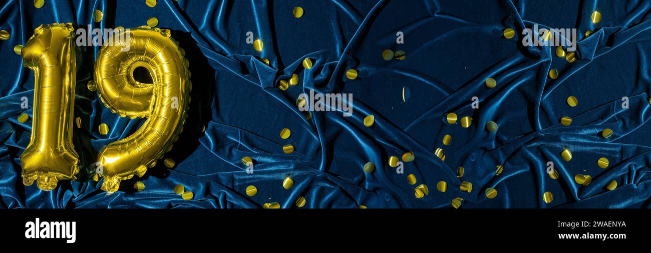 number nineteen, gold foil balloon number on a blue velvet background with confetti. Birthday ...
