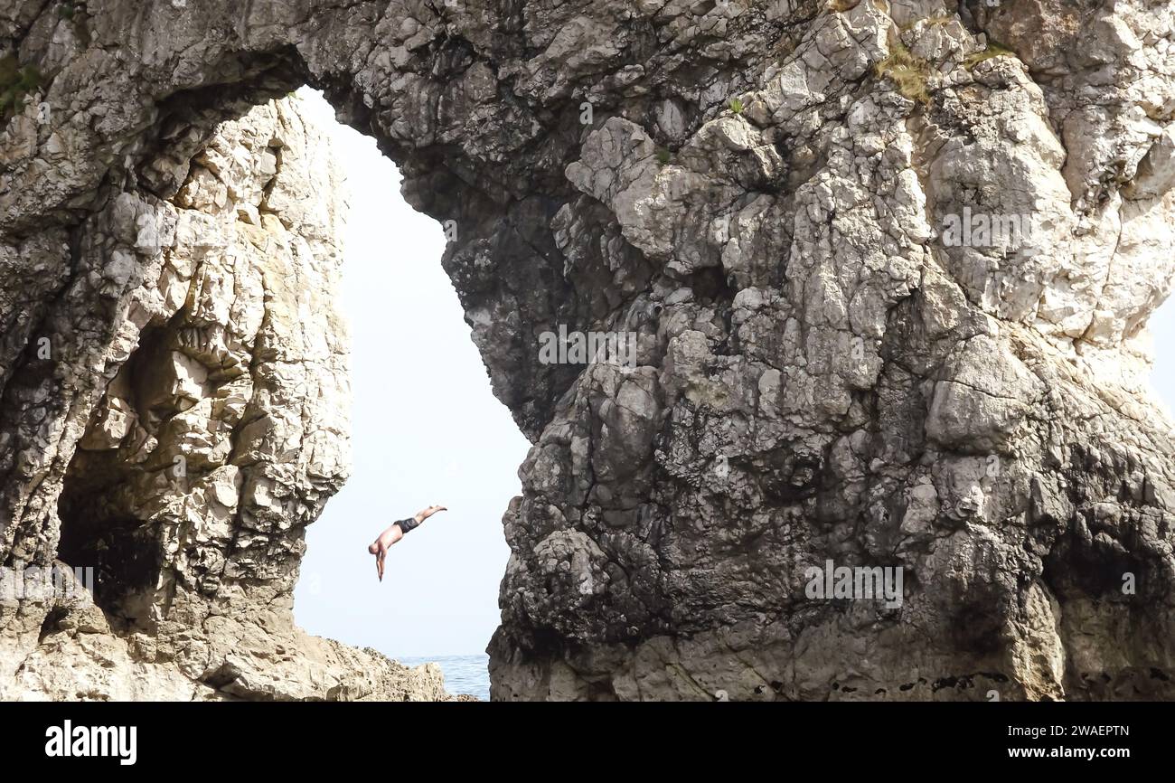 A young male mid-air in a daring jump from a cliff into the ocean below ...