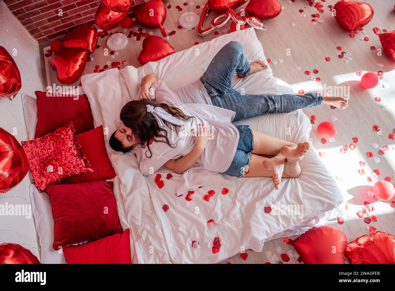 Couple In Love Hugs Tightly On White Bed Among Red Balloons Pillows