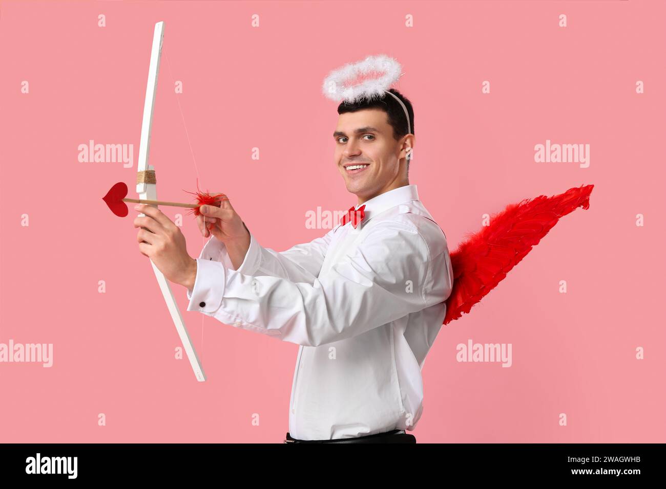 Young man dressed as Cupid with bow and arrow on pink background ...