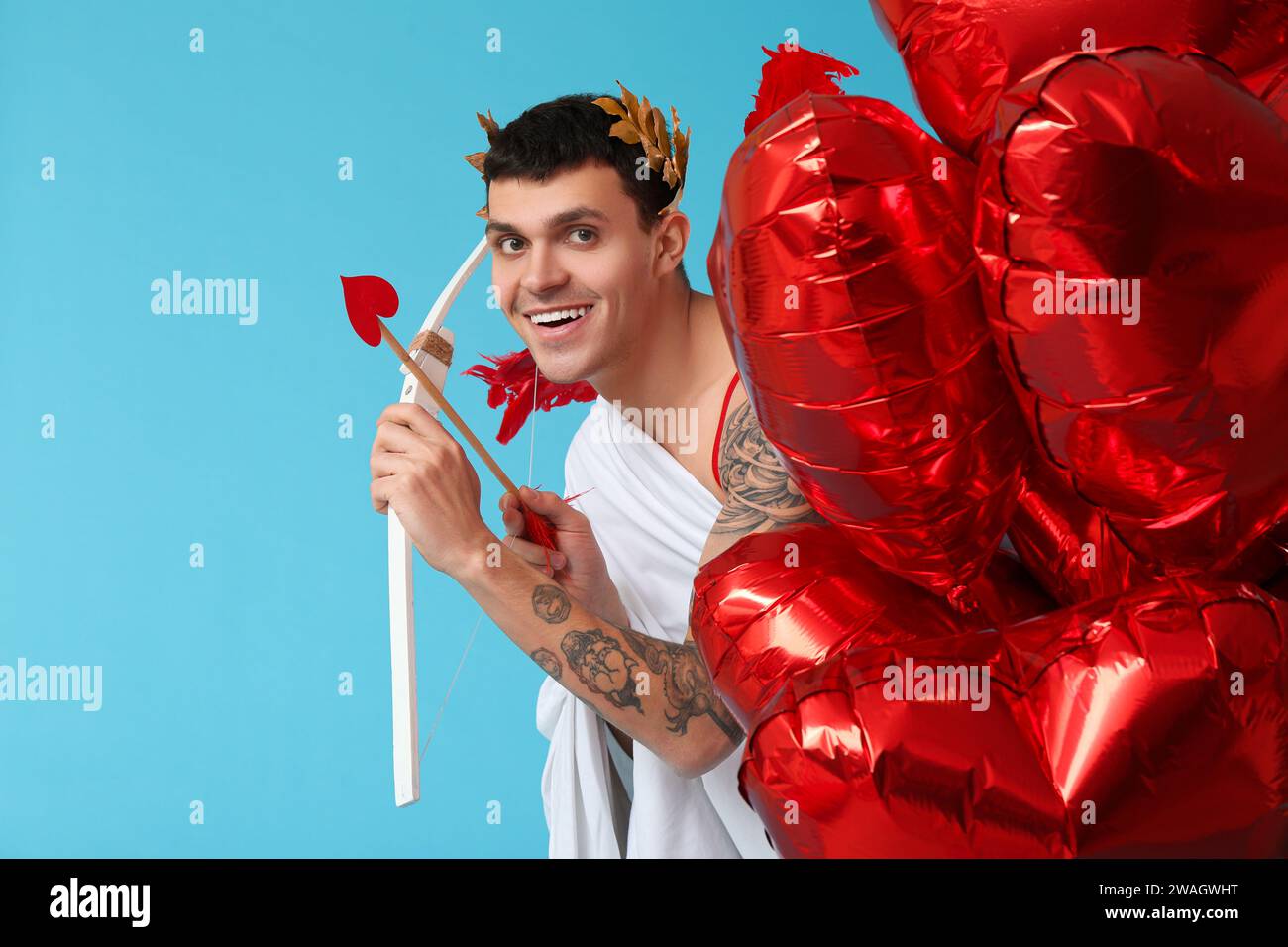 Young man dressed as Cupid with bow and balloons on blue background ...