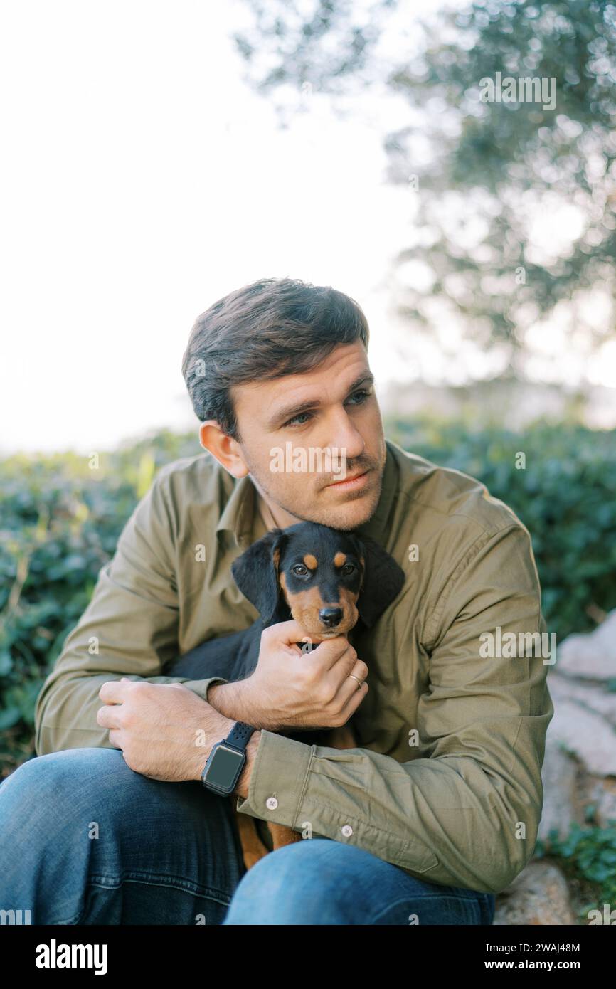 Pensive guy sits in a clearing with a puppy on his knees, put his chin ...