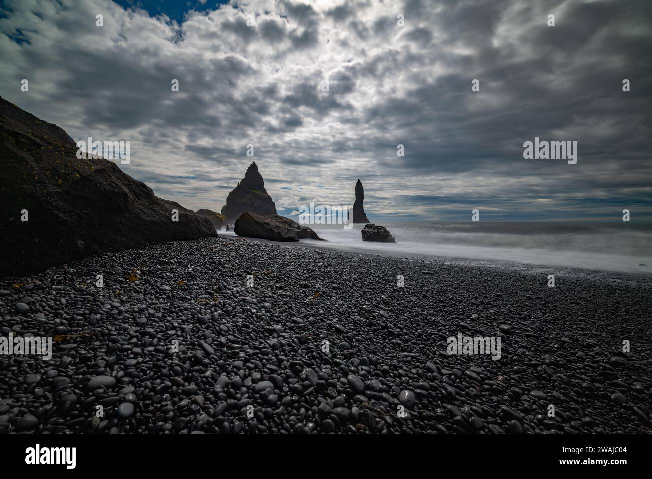 Jagged sea stacks rise from the misty ocean off the pebbled shore of a ...