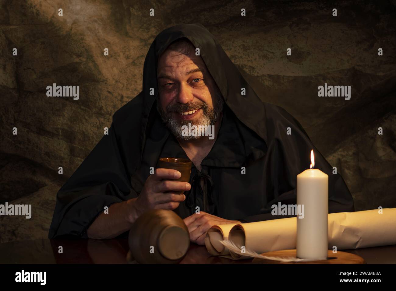 Cheerful monk in cassock drinking wine from a clay goblet sitting at a ...
