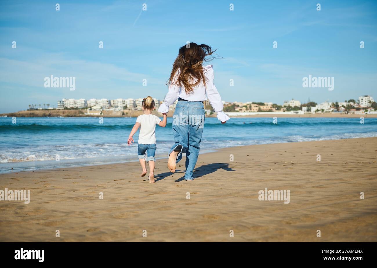 Rear view of a young mother and daughter holding hands, running ...