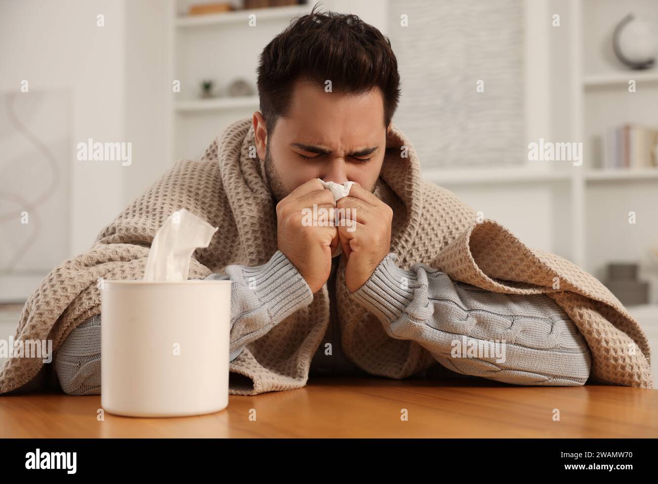 Sick man wrapped in blanket with tissue blowing nose at wooden table ...