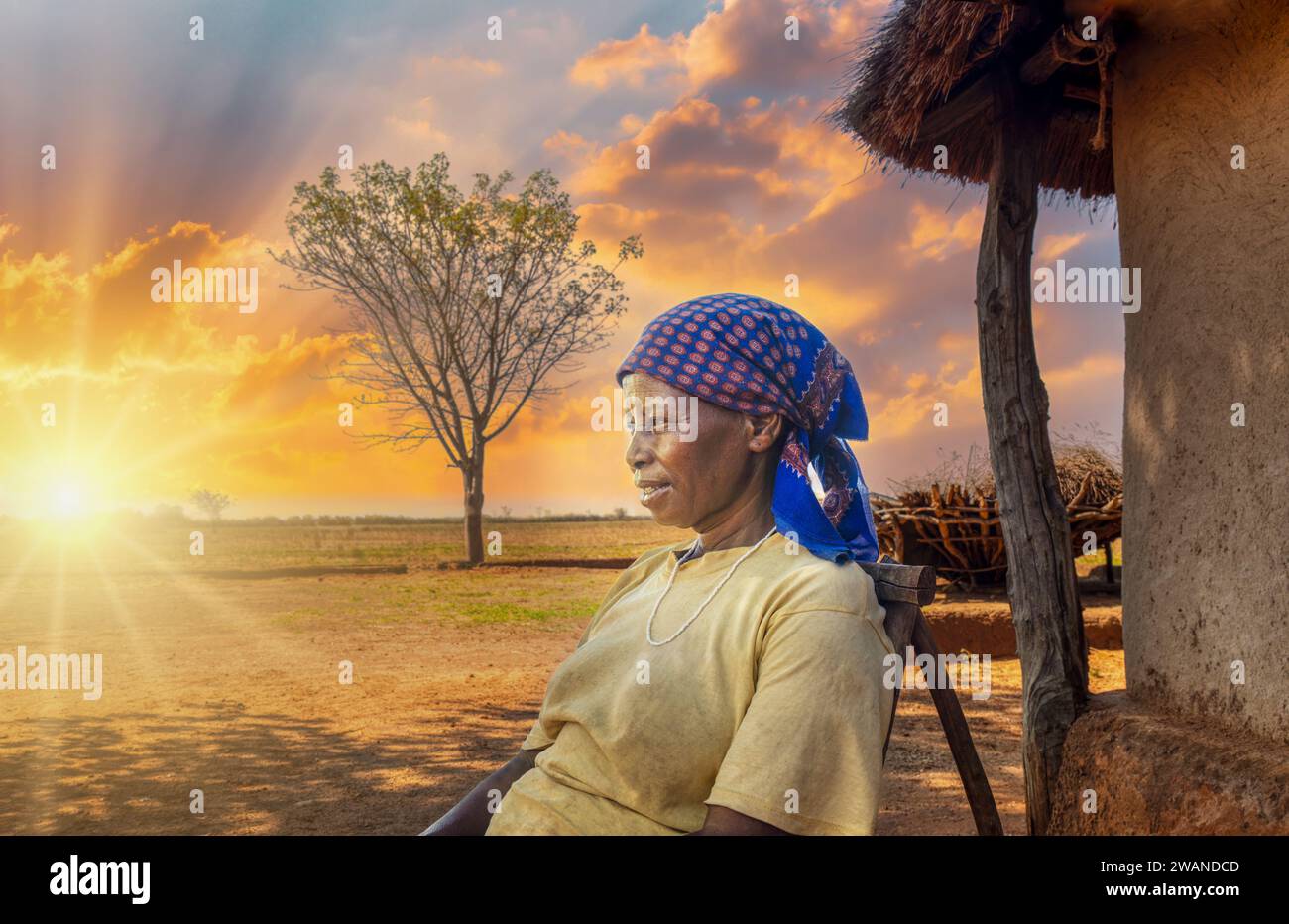 village african adult woman, sited on a chair in the yard at sunset in front of a thatched roof ...
