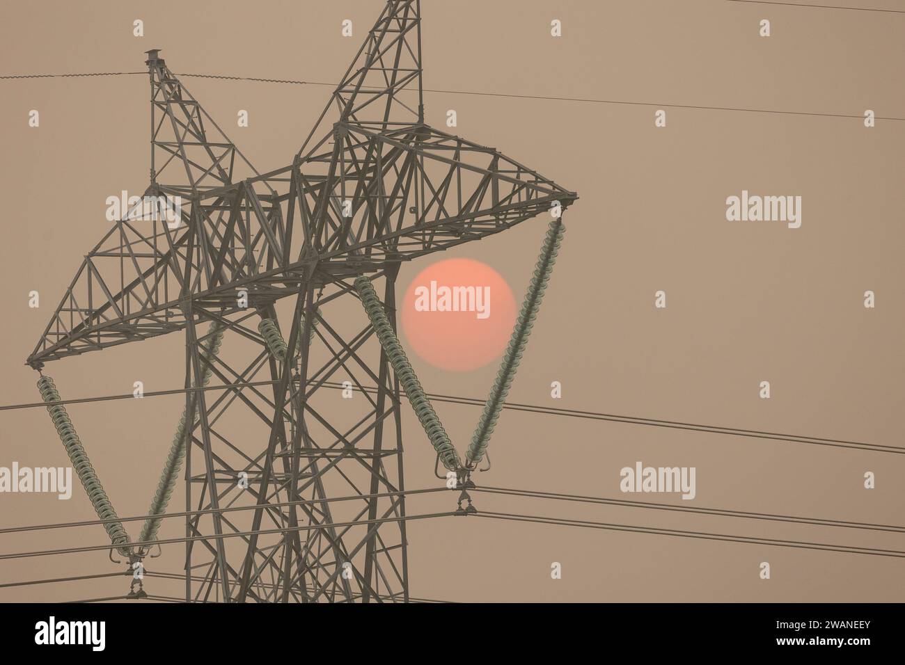 Hazy orange sun in smoky sky behind top of an electricity pylon ...