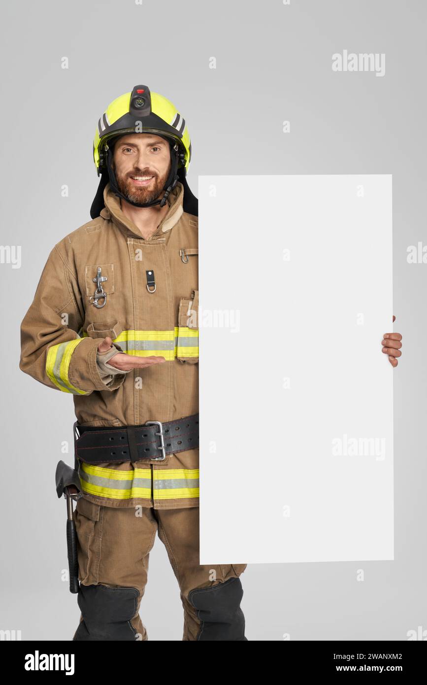 Happy firefighter in uniform standing with blank cardboard indoors ...