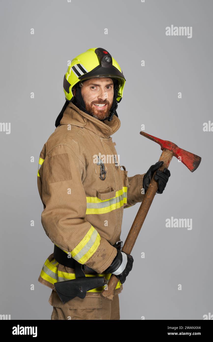 Smiling male firefighter in full gear holding big axe for work. Side ...