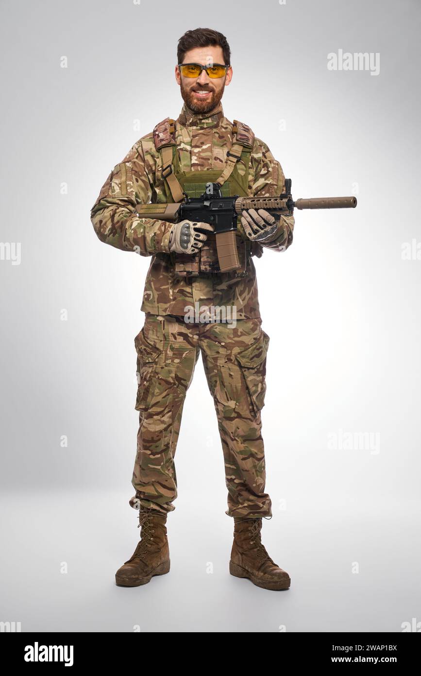 Smiling armed soldier posing with assault rifle, looking camera indoors ...