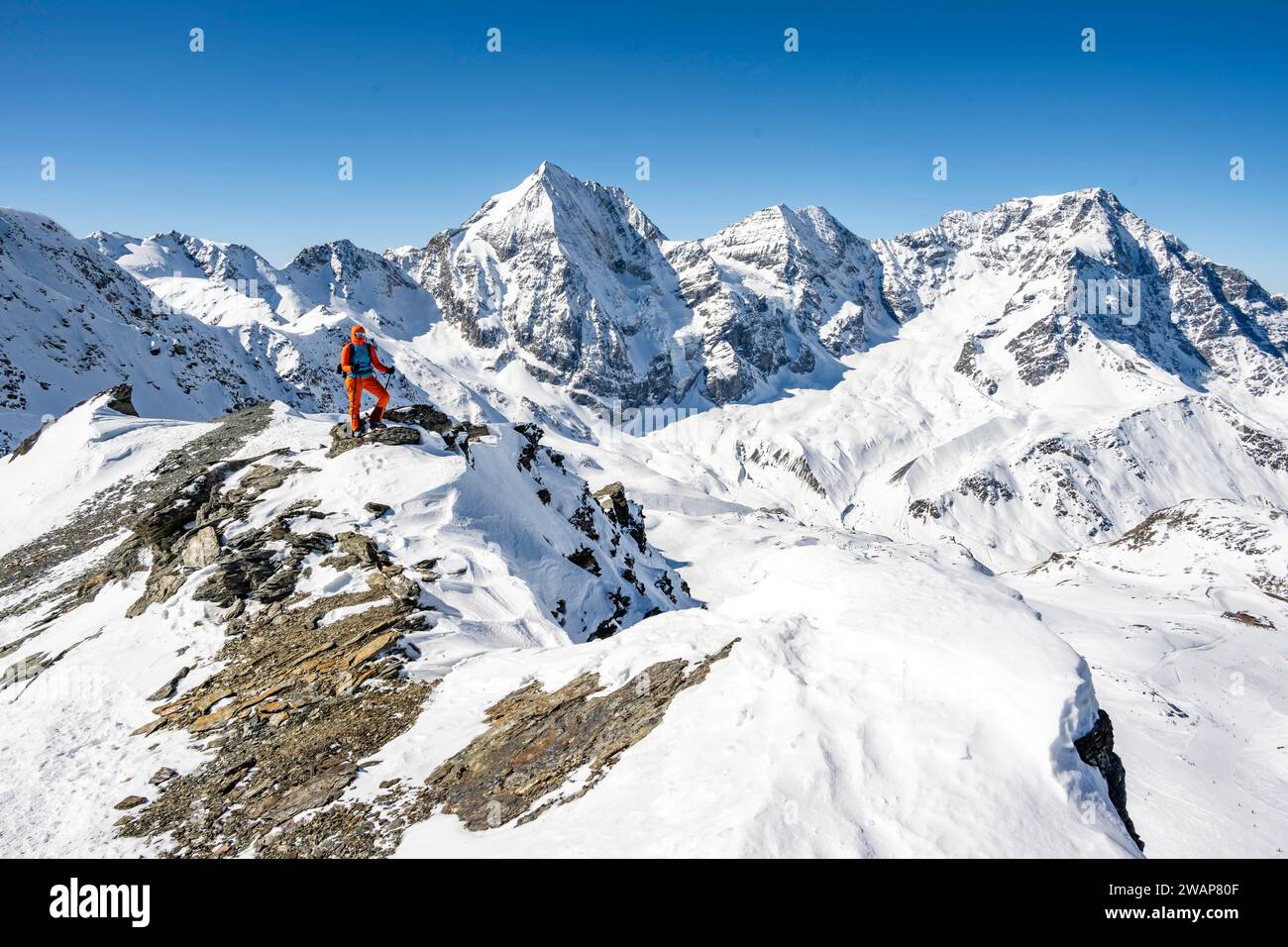 Mountaineer standing on a ledge in front of snow-covered mountains ...