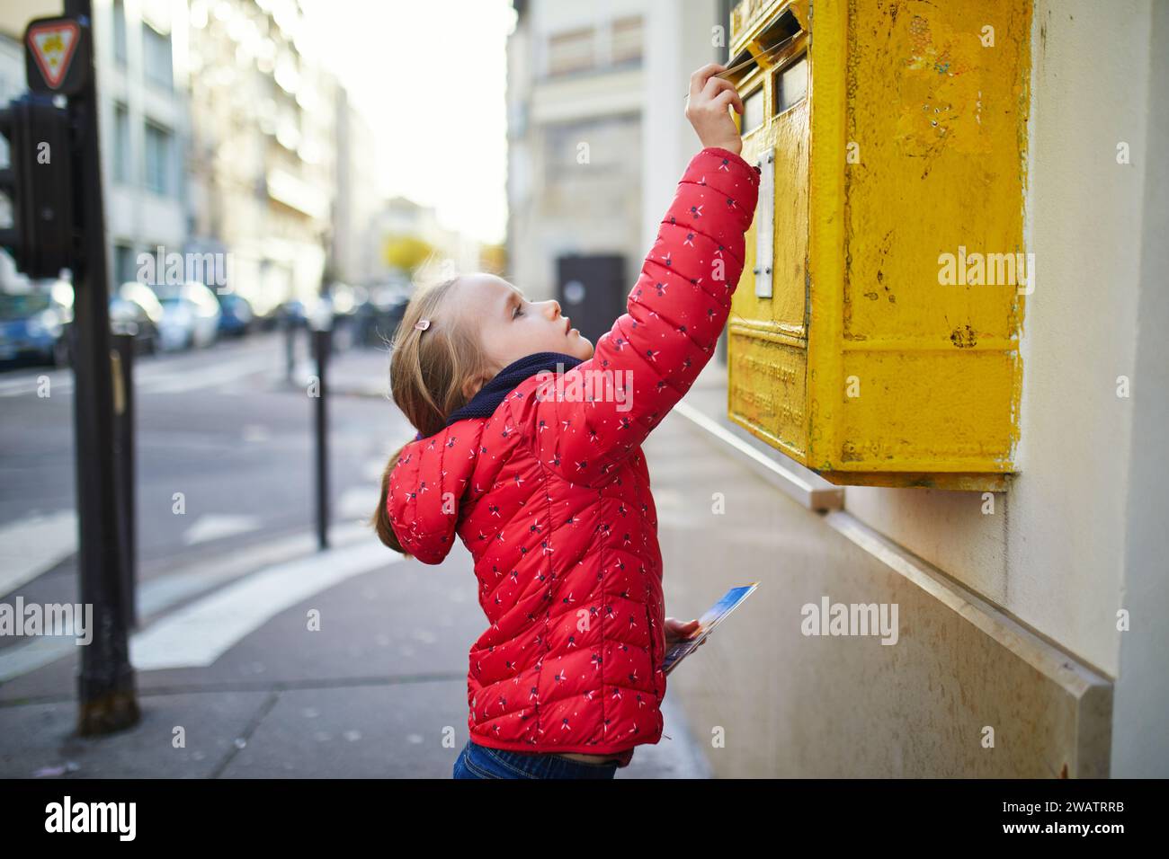 Adorable preschooler girl putting letter in yellow post box on a street ...