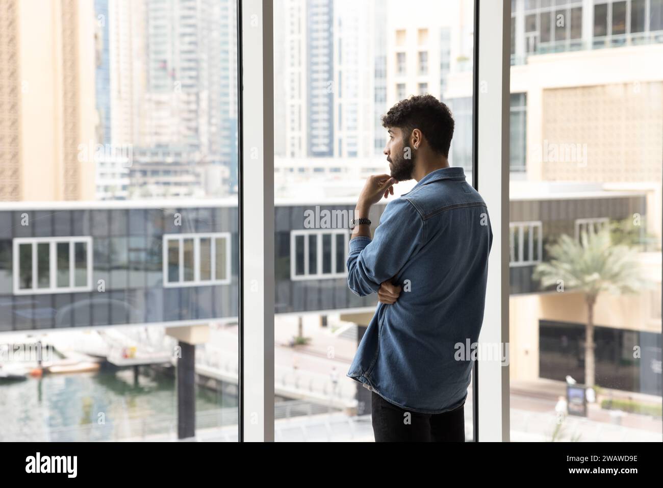 Thoughtful serious handsome young Arab man standing at large window ...
