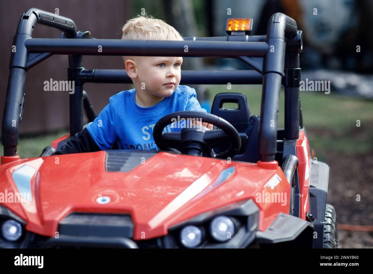 Happy blond little boy in a big red toy battery powered car outdoors ...