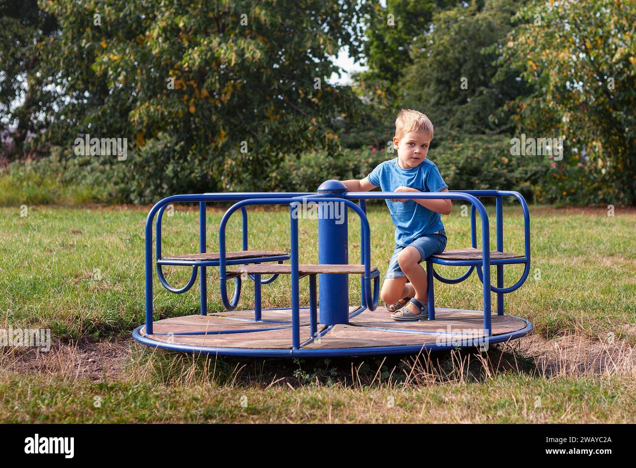 Cute Caucasian boy in blue clothes swings on outdoor park carousel against autumn tree foliage ...