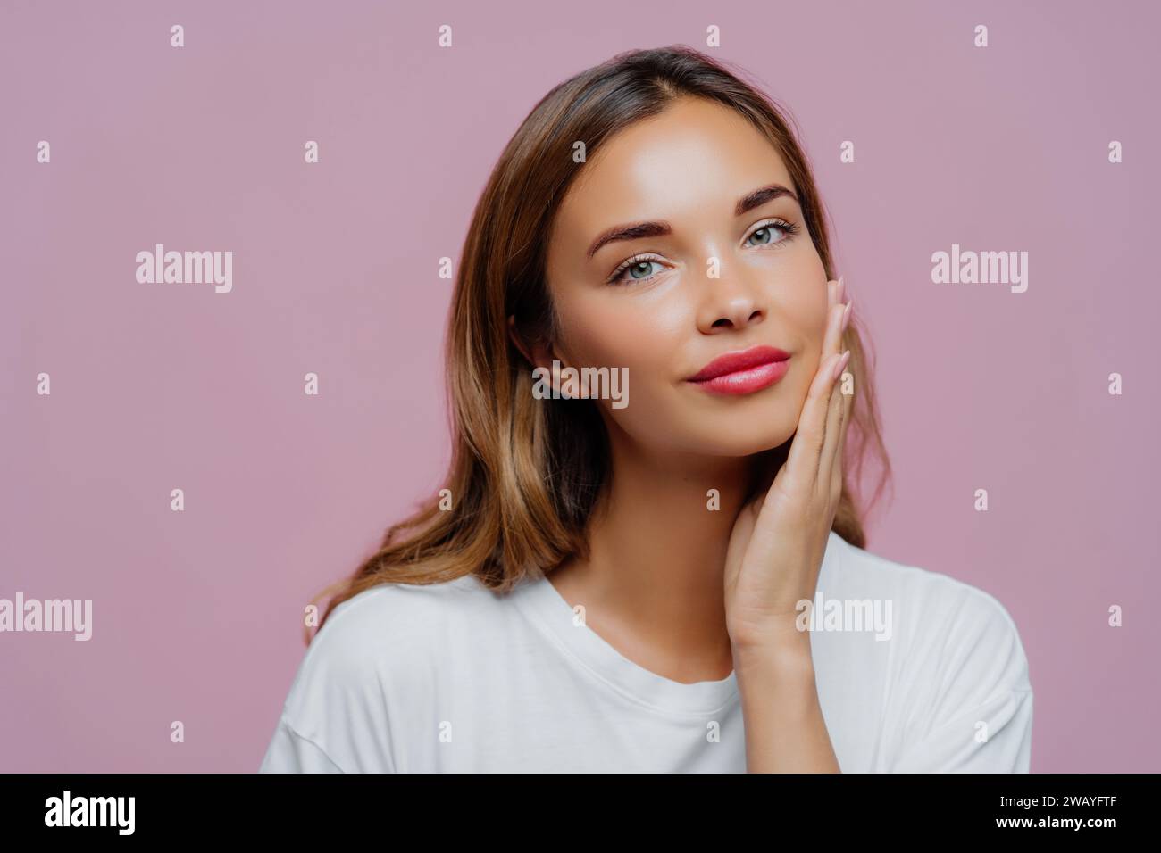 Contemplative woman with hand on cheek, white tee, pink background ...