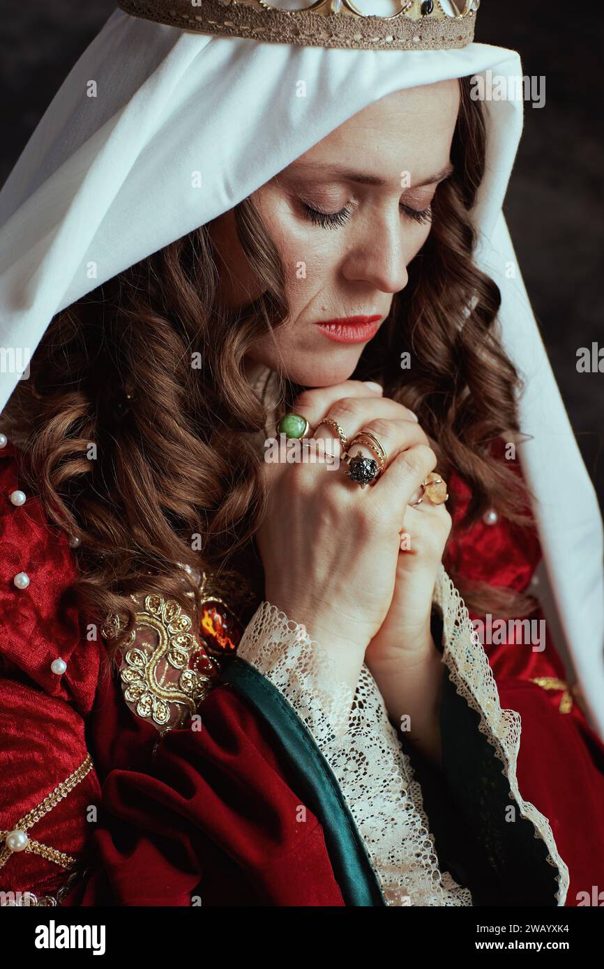medieval queen in red dress with veil and crown praying on dark gray ...