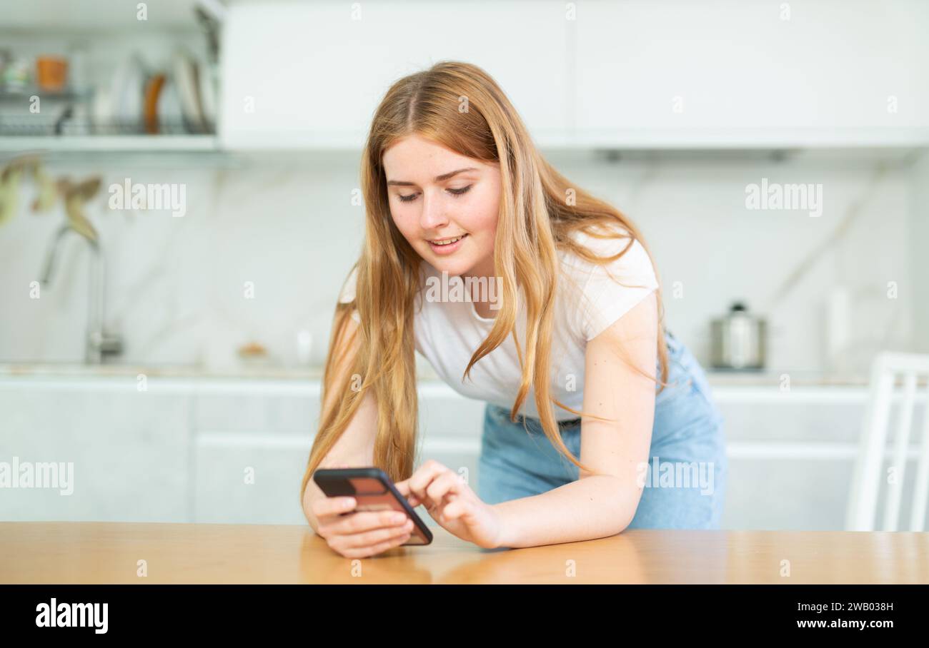 bored young woman stands in kitchen leans on countertop and surfs ...