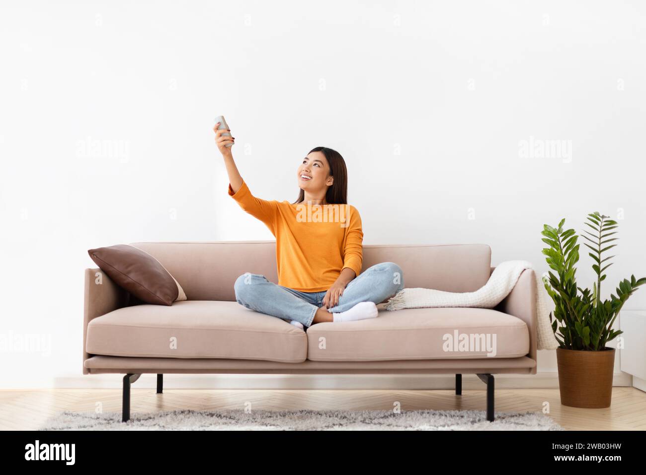 Relaxed millennial korean woman sitting on couch, turning on AC Stock ...