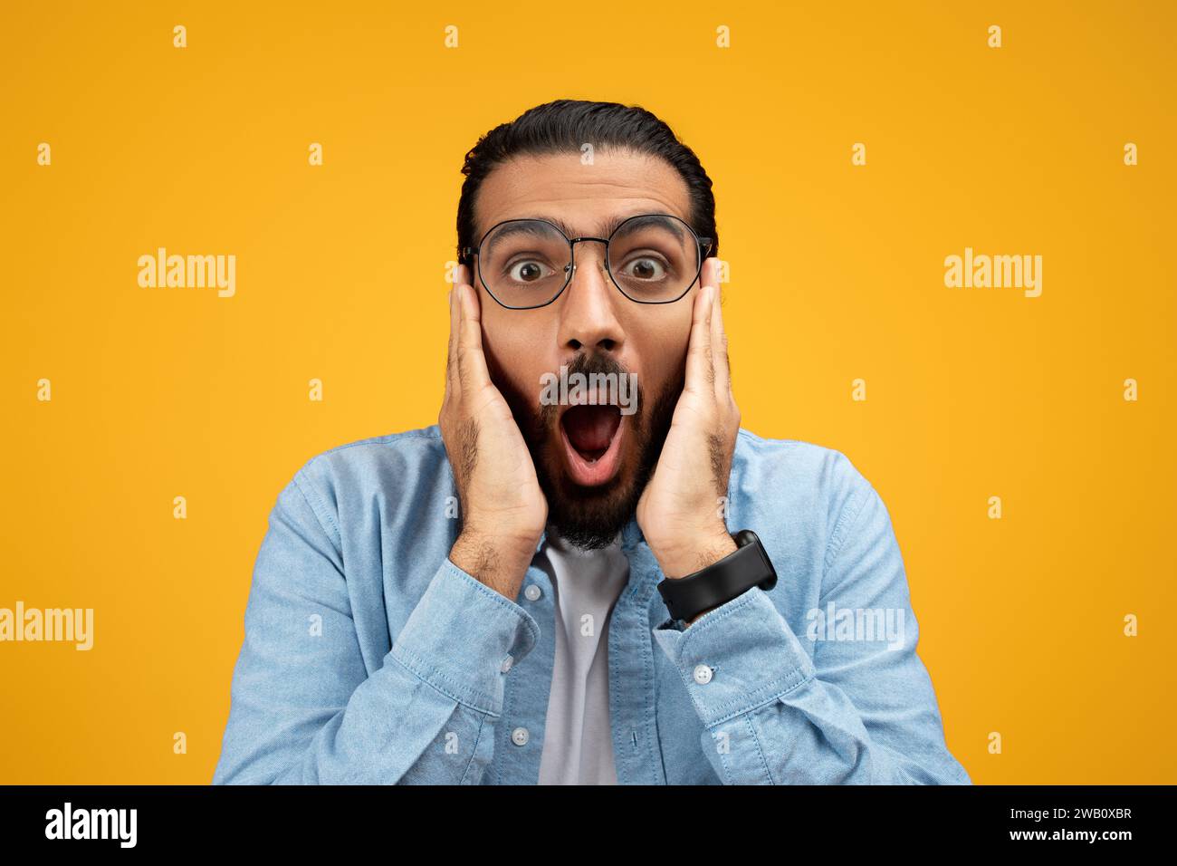 Shocked man in a blue shirt with hands on cheeks and mouth wide open ...