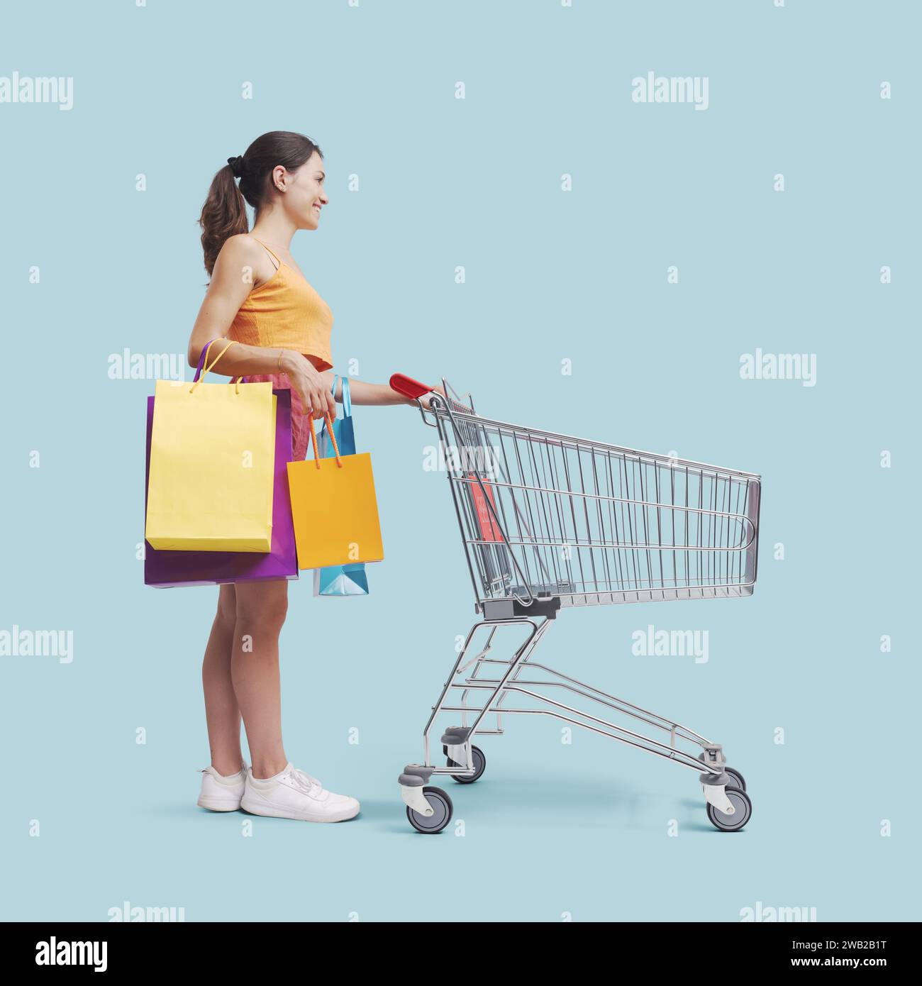 Young woman pushing an empty shopping cart, holding shopping bags and ...