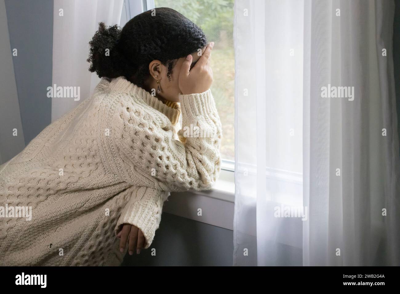Biracial teen girl by window with hands over face looking depressed ...