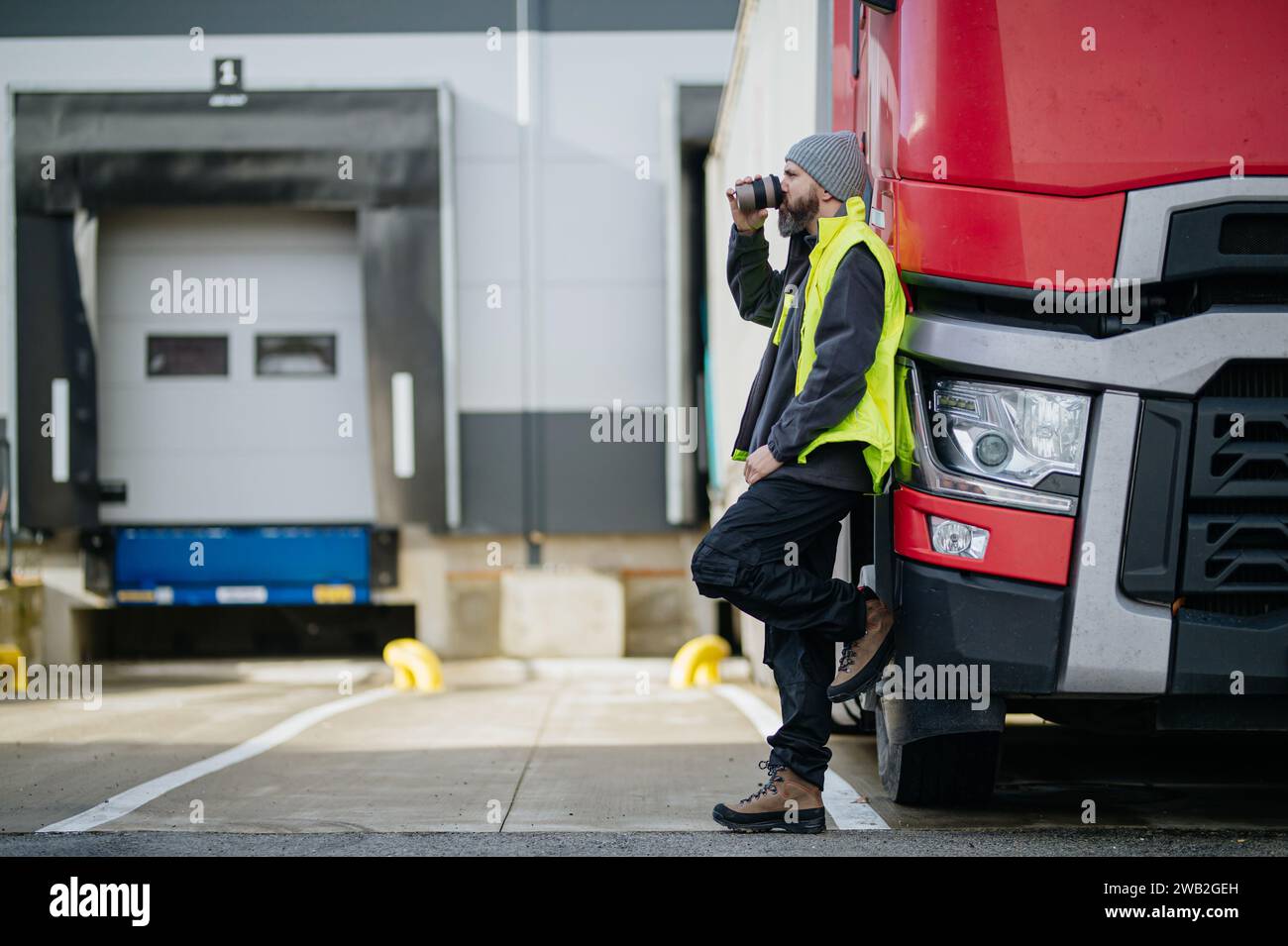 Truck driver leaning against red truck and drinking coffee, waiting for ...