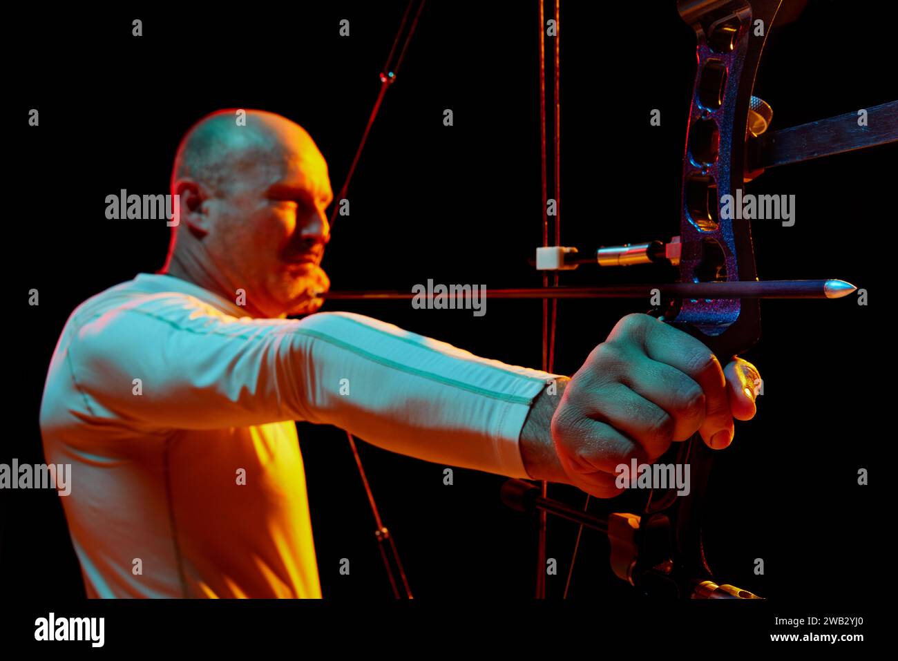 Portrait of serious man, archer aiming with bow on target against black studio background in ...