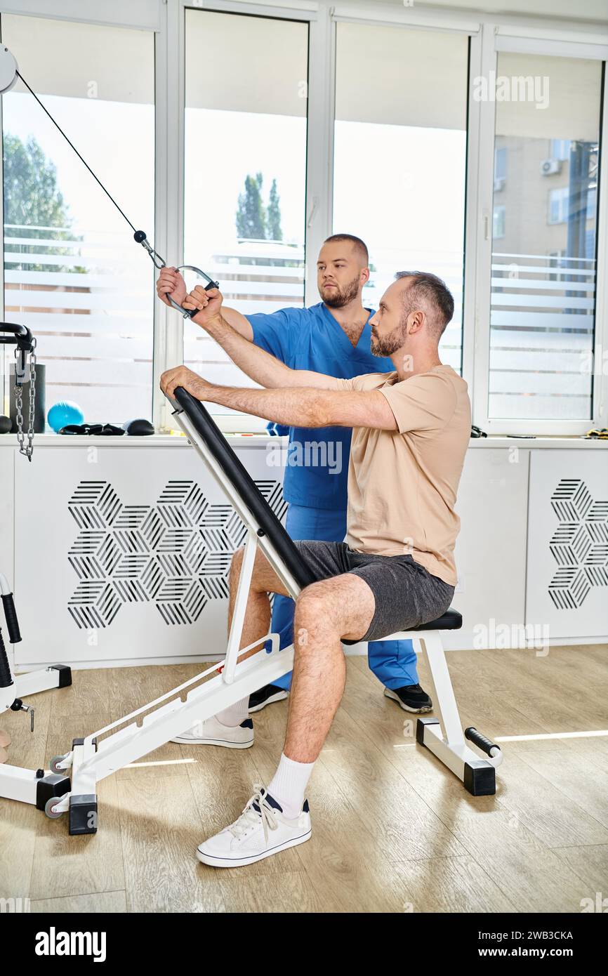 skilled rehabilitologist in blue uniform assisting man during recovery training in kinesio ...