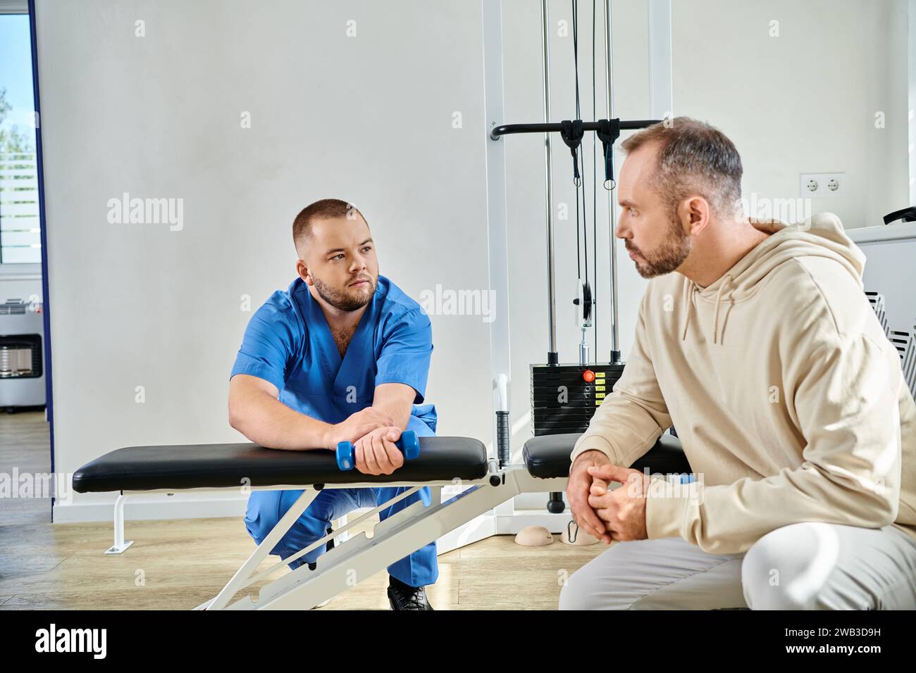male patient looking at rehabilitologist showing exercise with dumbbell ...